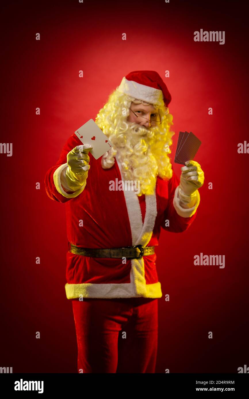 Santa Claus holds playing cards in his hands and poses on a dark red ...