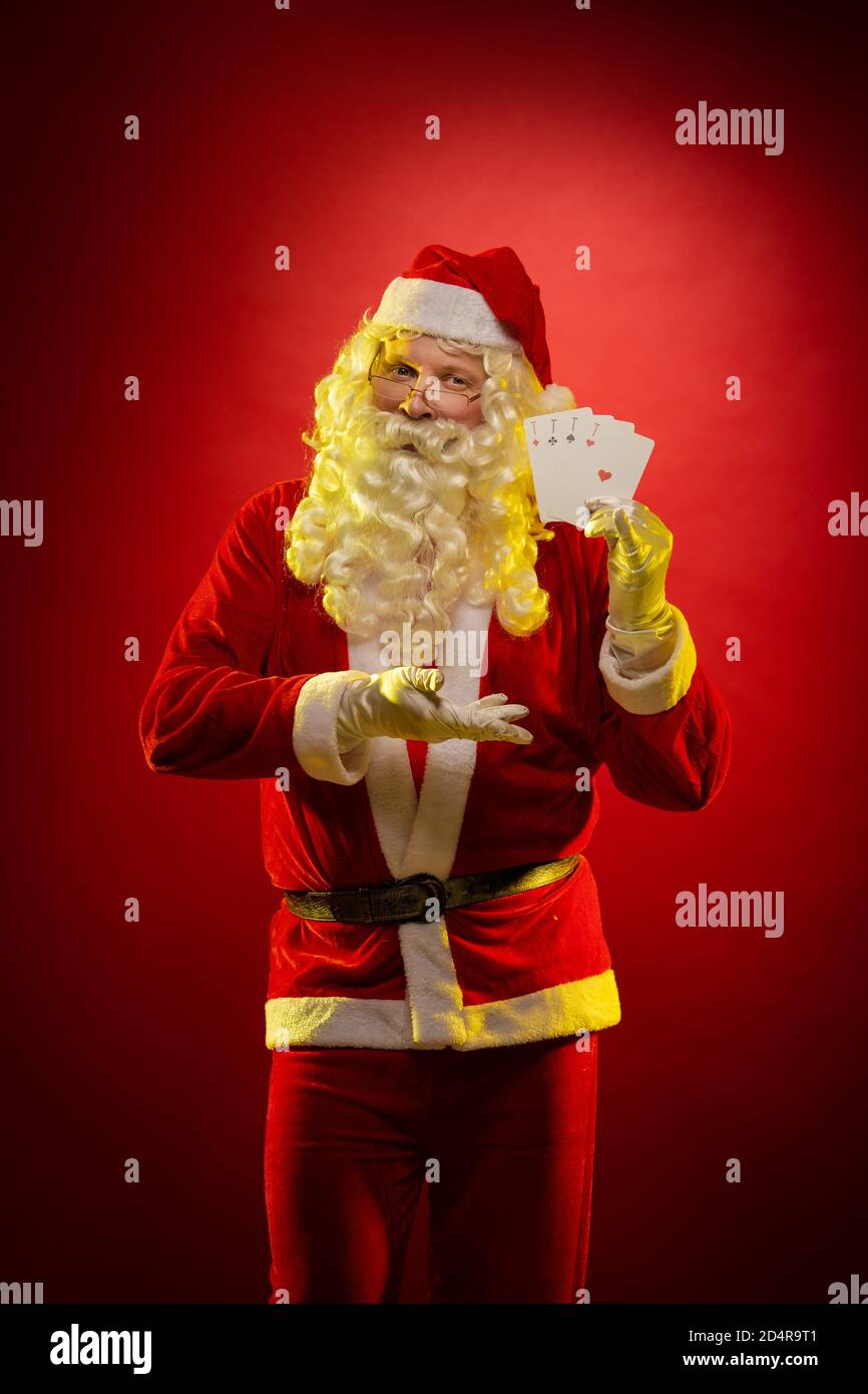 Santa Claus holds playing cards in his hands and poses on a dark red ...