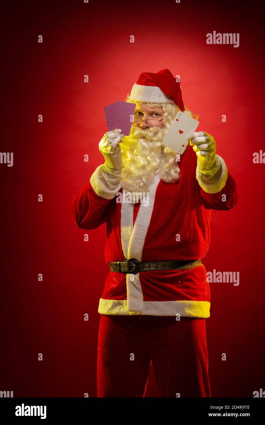 Santa Claus holds playing cards in his hands and poses on a dark red ...