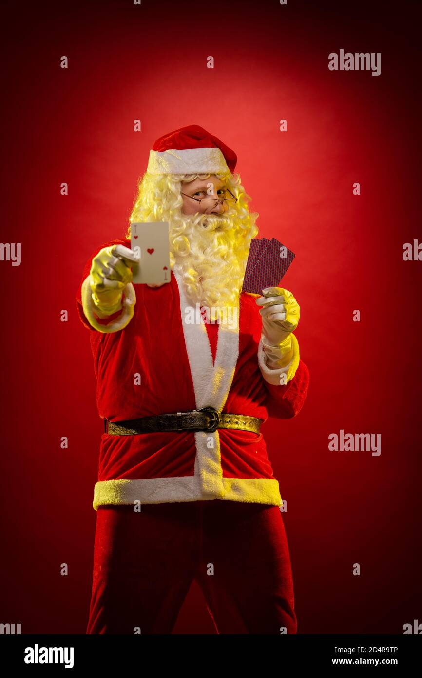 Santa Claus holds playing cards in his hands and poses on a dark red ...