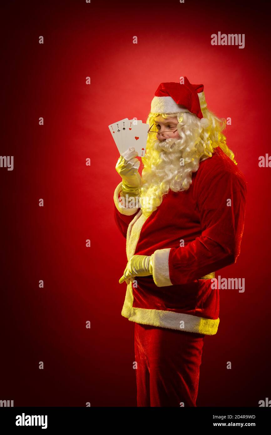 Santa Claus holds playing cards in his hands and poses on a dark red ...