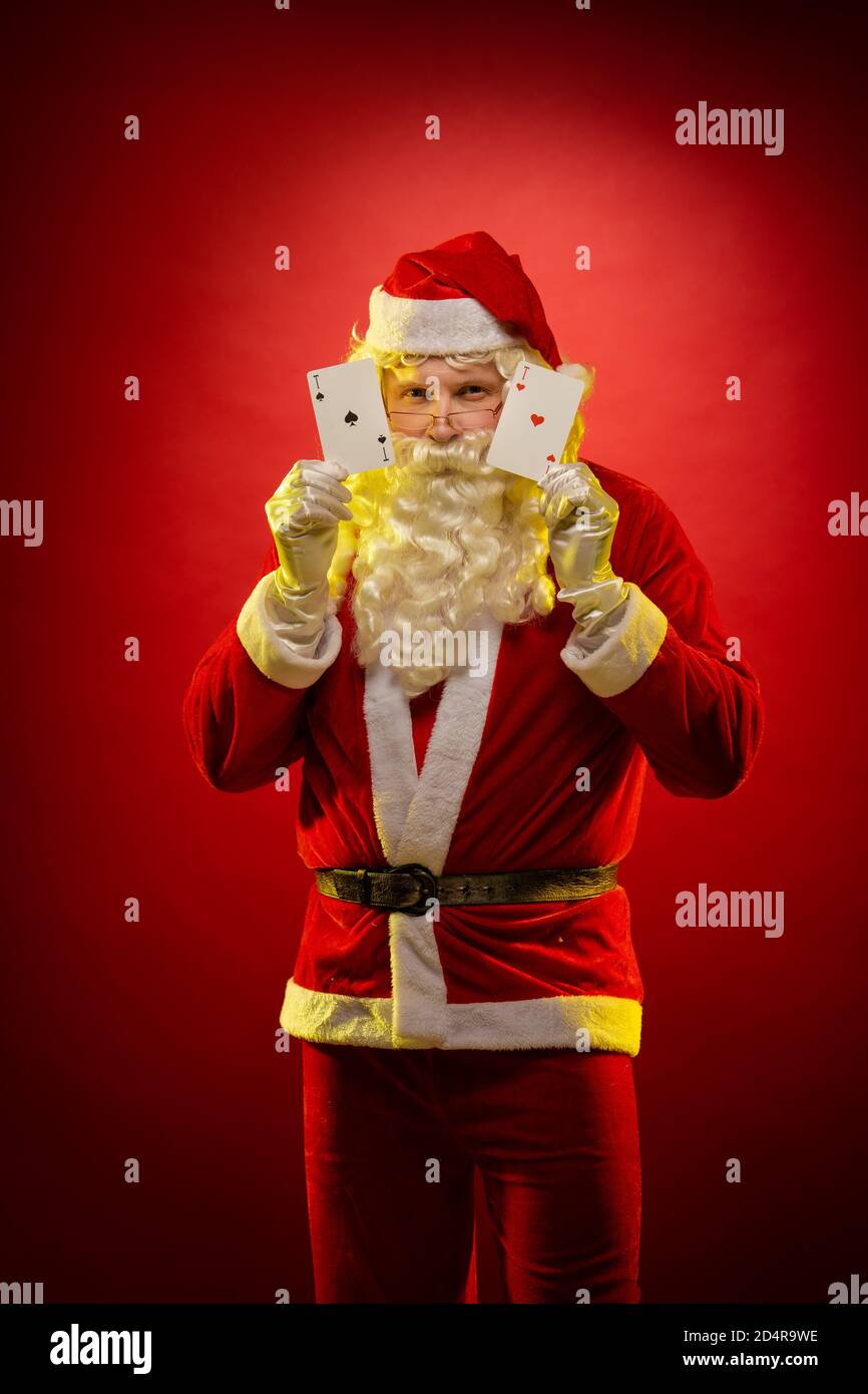 Santa Claus holds playing cards in his hands and poses on a dark red ...