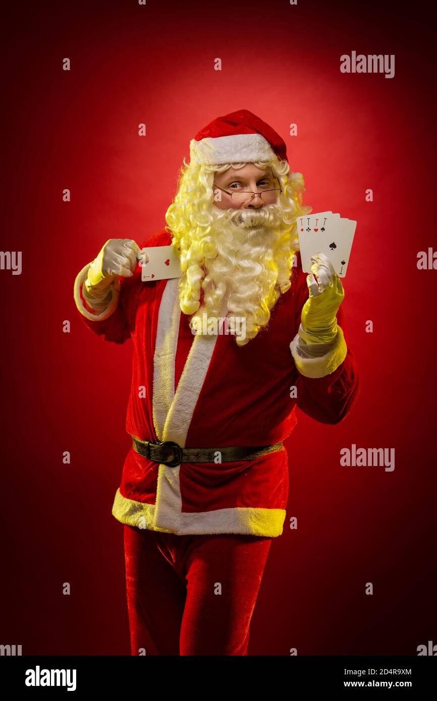 Santa Claus holds playing cards in his hands and poses on a dark red ...