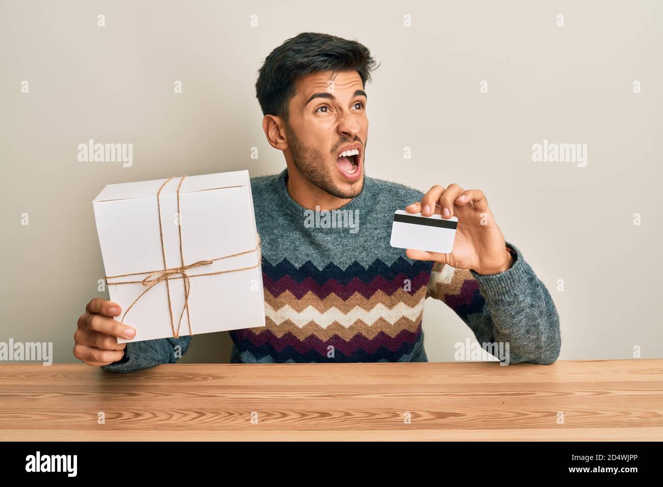 Young handsome man holding gift and credit card angry and mad screaming ...