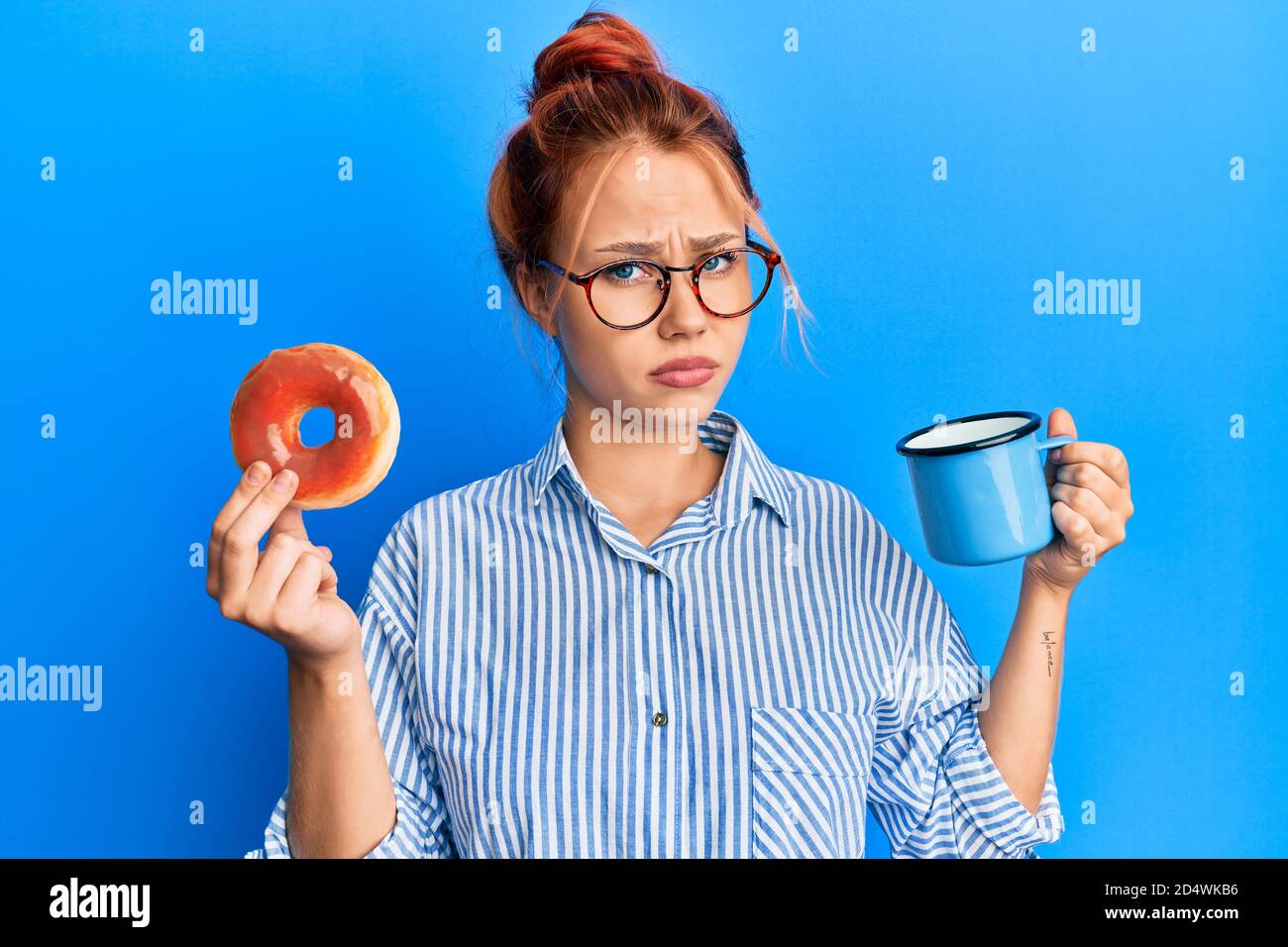 Young redhead woman eating breakfast holding chocolate donut and coffee ...