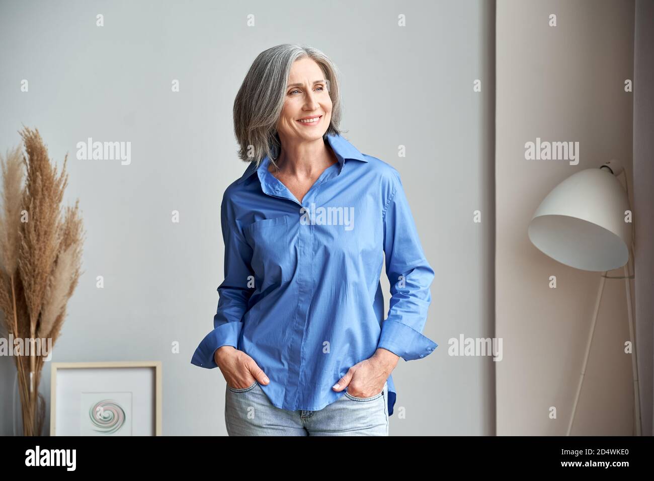 Smiling confident mature older woman standing indoors looking at window ...