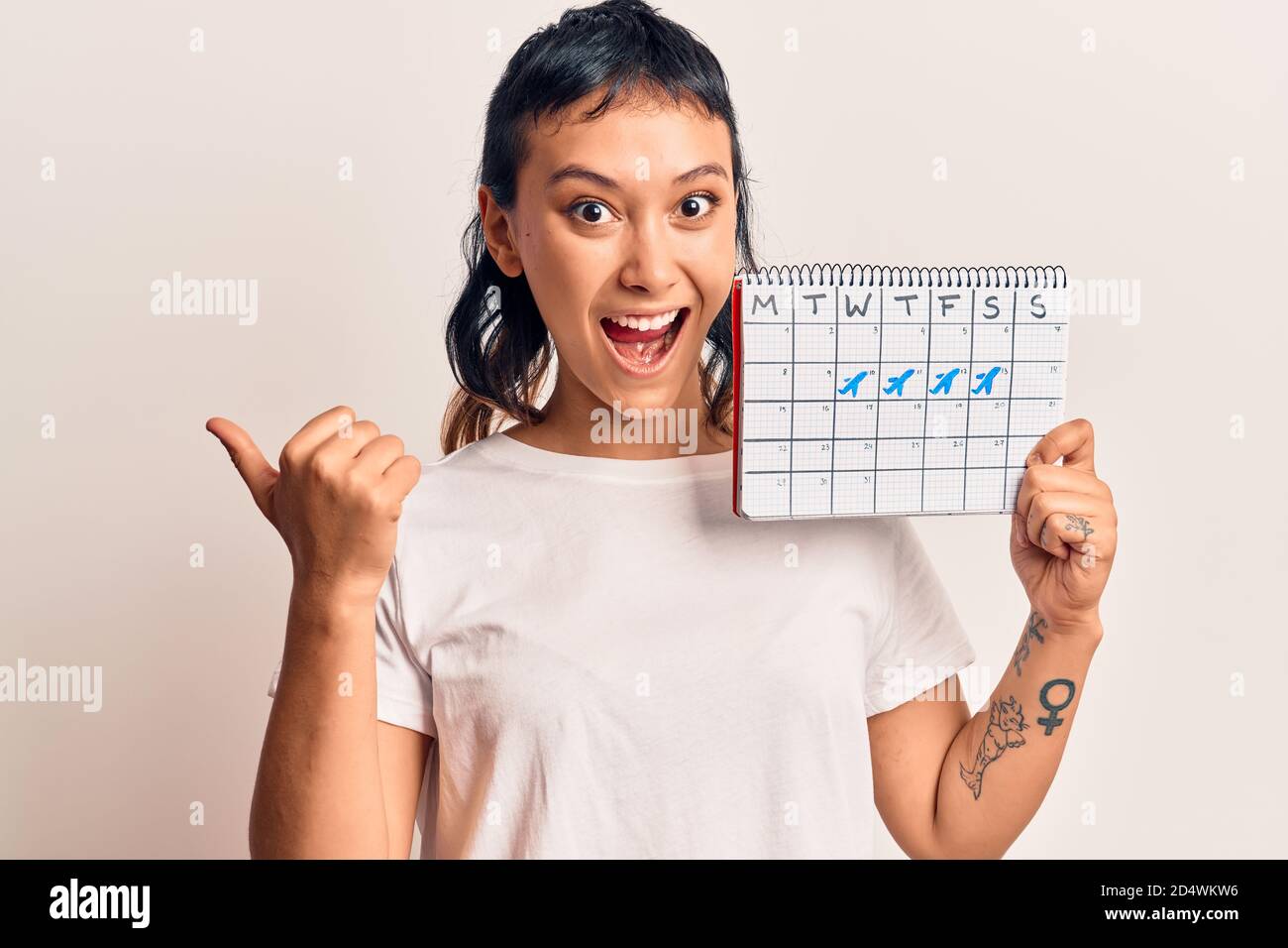 Young woman holding travel calendar pointing thumb up to the side ...