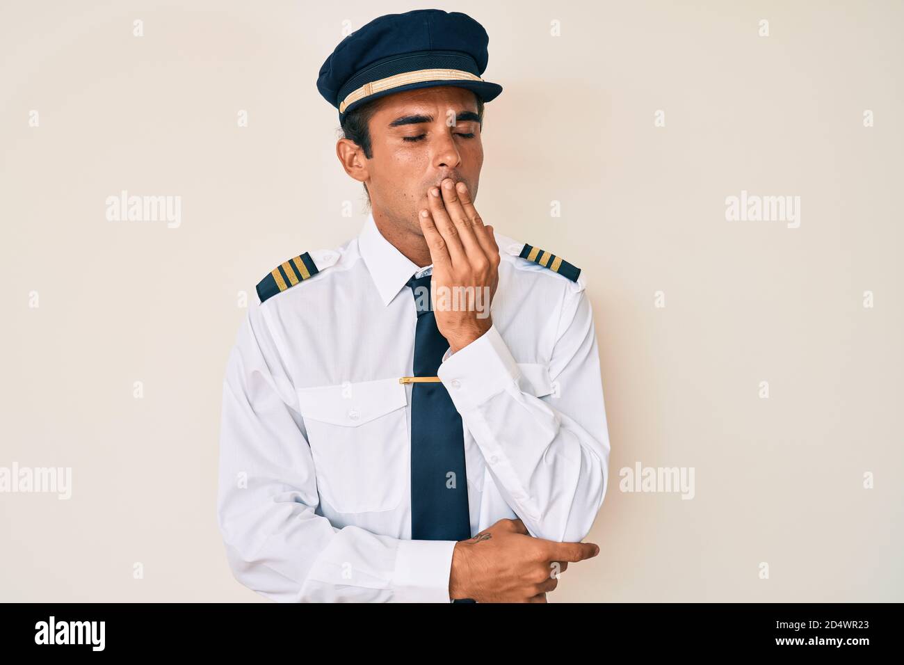 Young hispanic man wearing airplane pilot uniform bored yawning tired ...