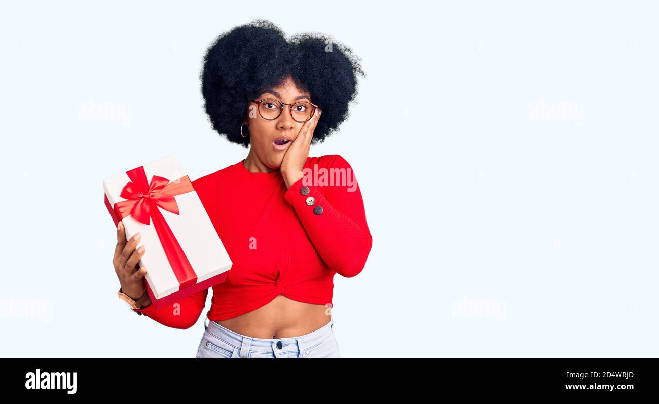 Young african american girl holding gift afraid and shocked, surprise ...