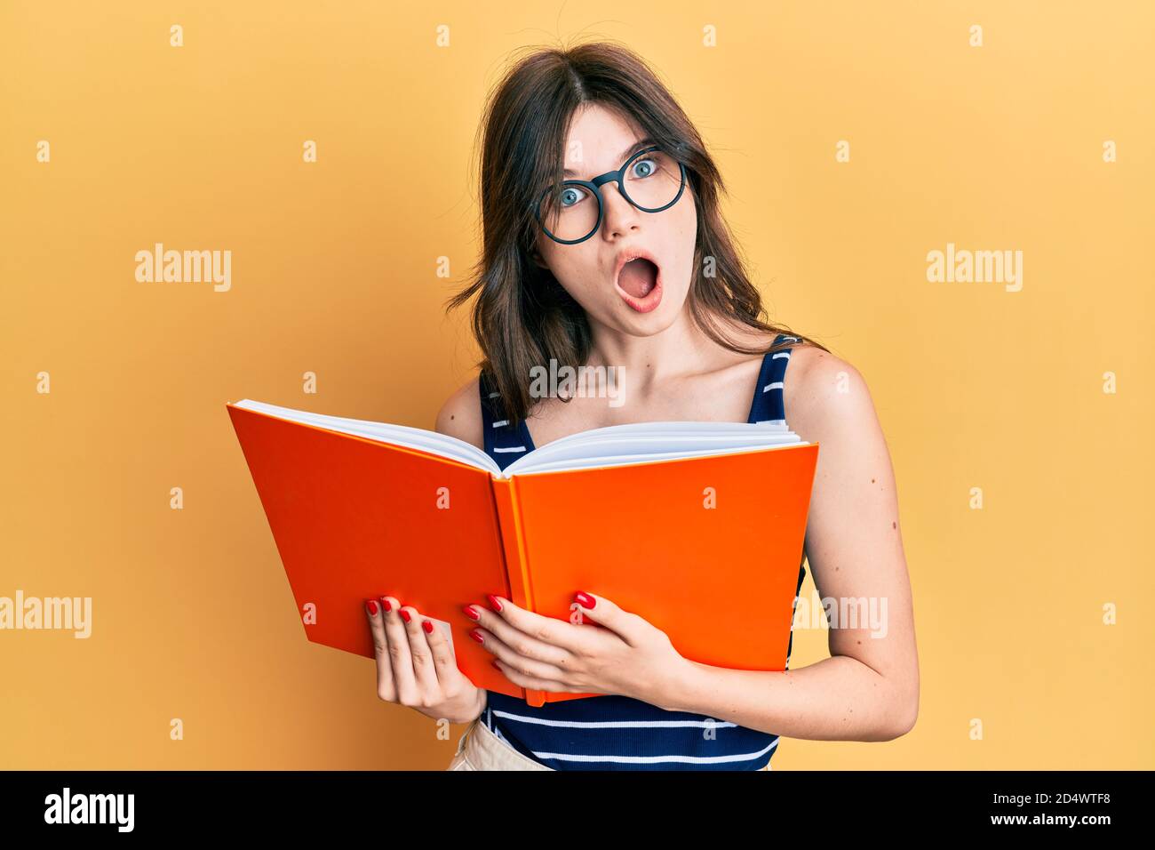 Young beautiful caucasian girl reading a book wearing glasses in shock ...