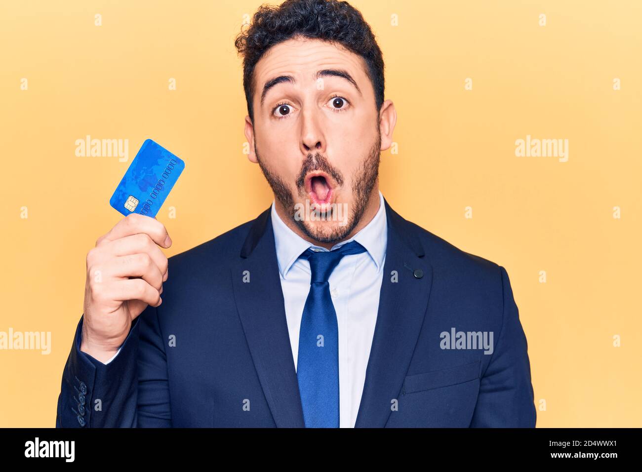 Young hispanic man wearing suit holding credit card scared and amazed ...