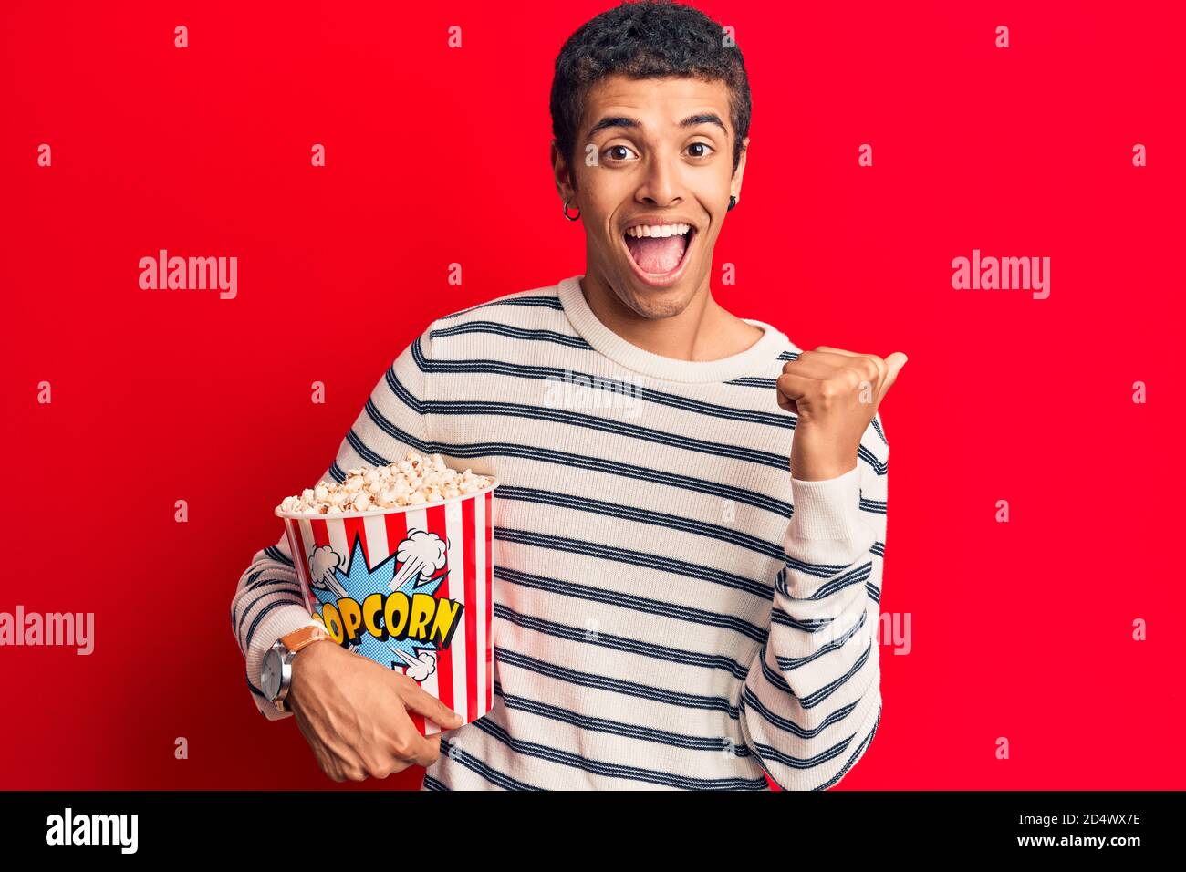 Young african amercian man holding popcorn pointing thumb up to the side smiling happy with open ...