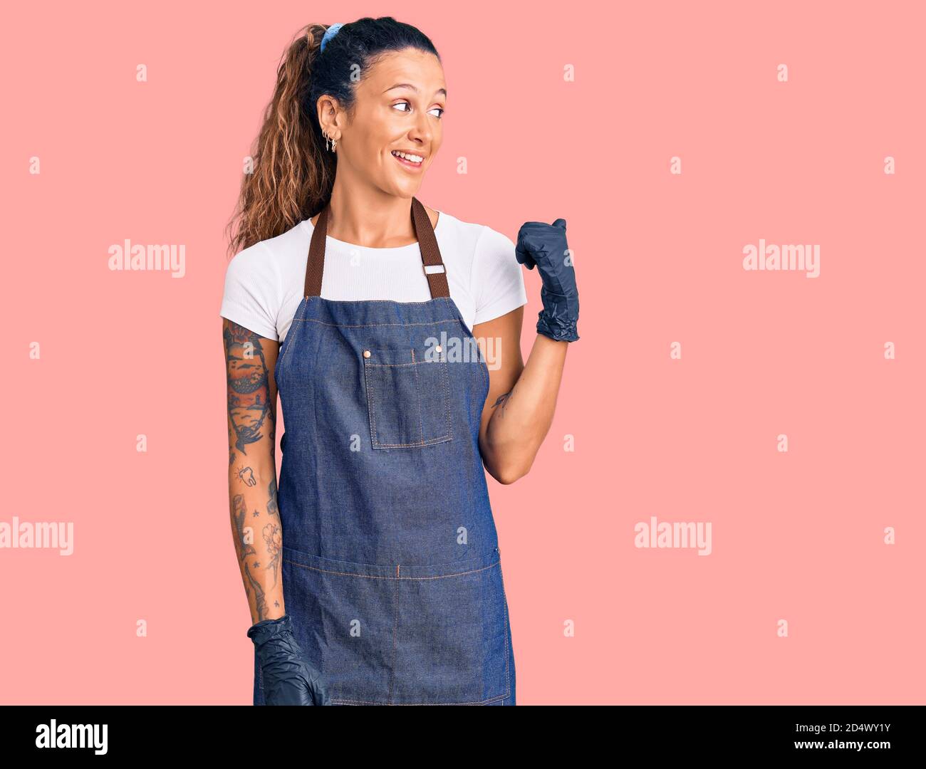 Young hispanic woman with tattoo wearing barber apron and gloves ...