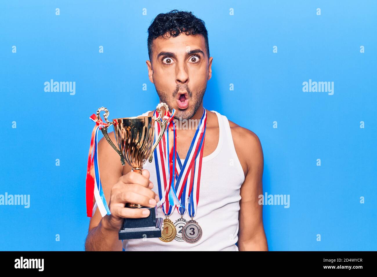 Young hispanic man wearing winner medals holding trophy scared and ...