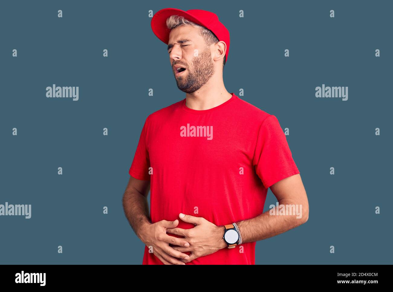 Young handsome blond man wearing t-shirt and cap with hand on stomach ...