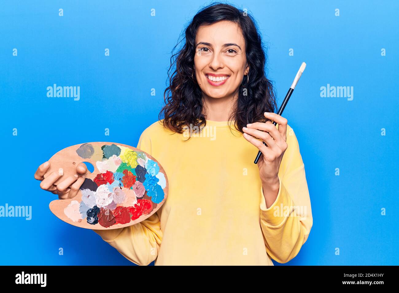 Young beautiful hispanic woman holding paintbrush and palette looking ...