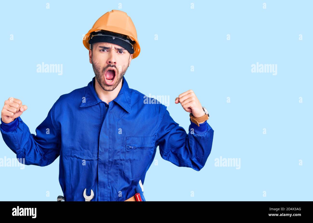 Young hispanic man wearing worker uniform angry and mad raising fists ...