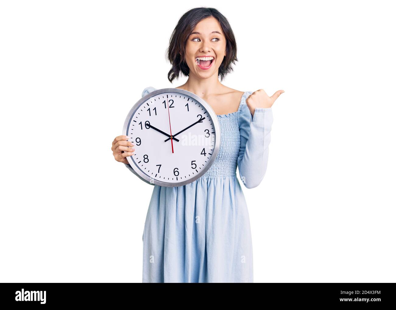 Young beautiful girl holding big clock pointing thumb up to the side ...