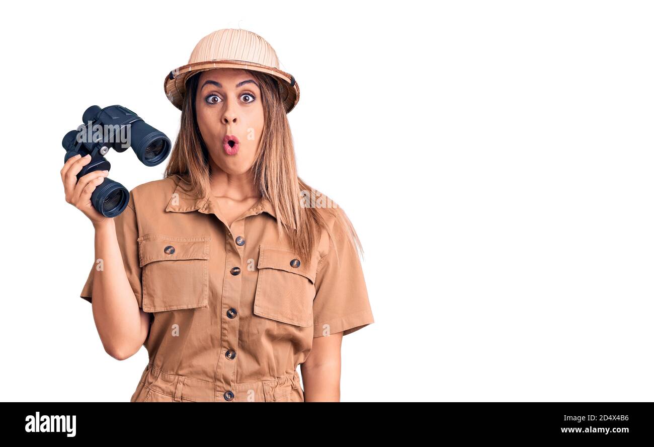 Young beautiful woman wearing explorer hat holding binoculars scared ...