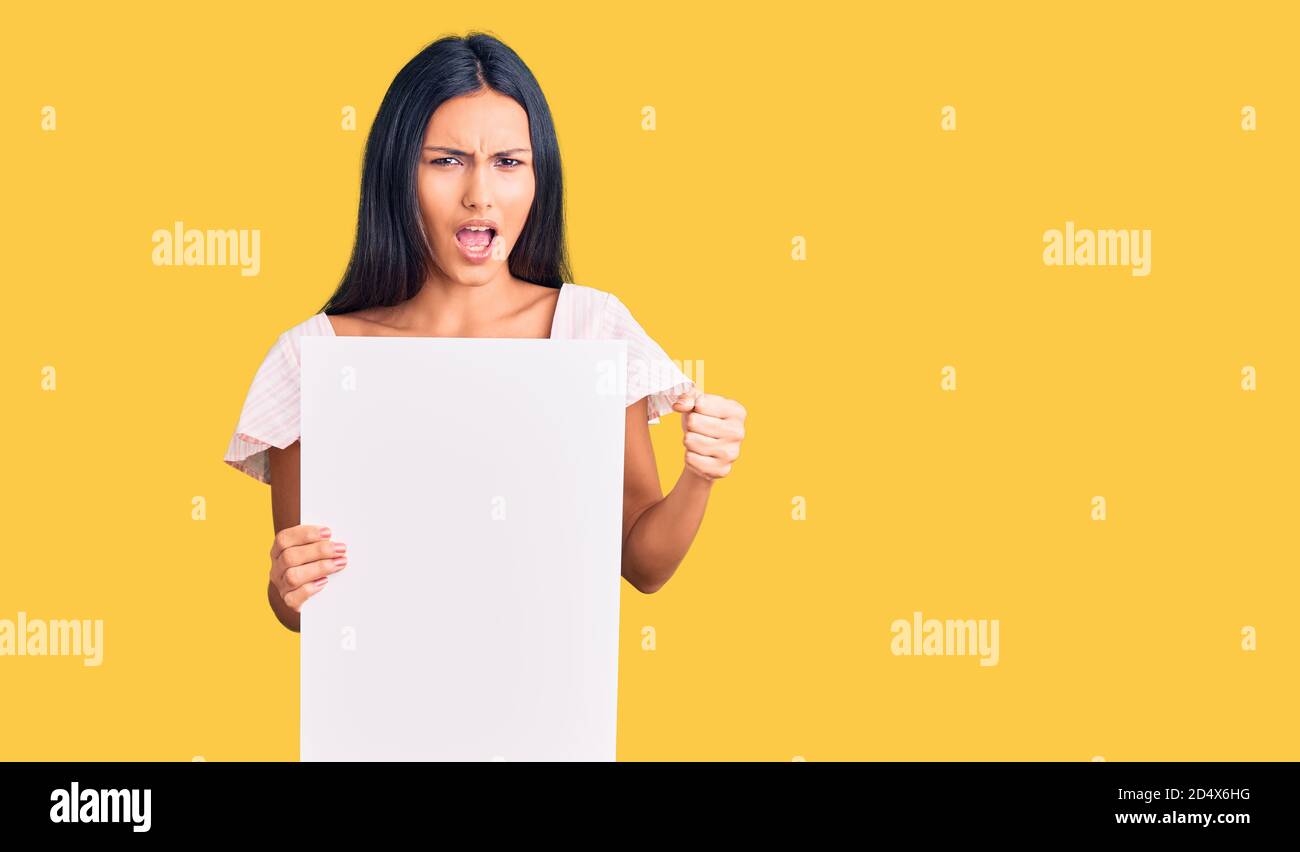 Young beautiful latin girl holding blank empty banner annoyed and ...