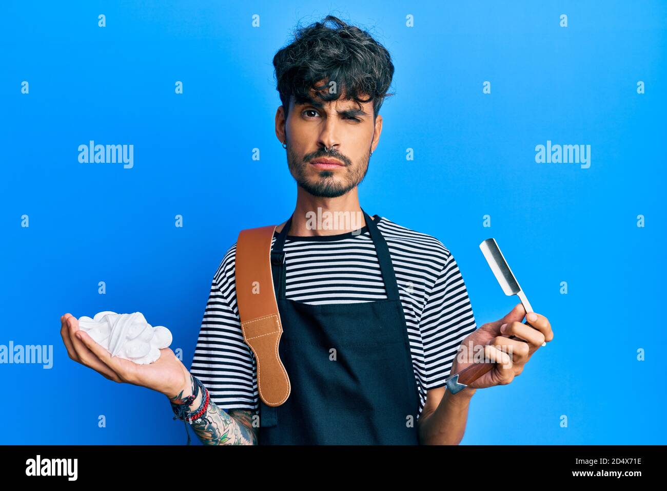 Young hispanic man wearing barber apron holding razor and foam skeptic ...