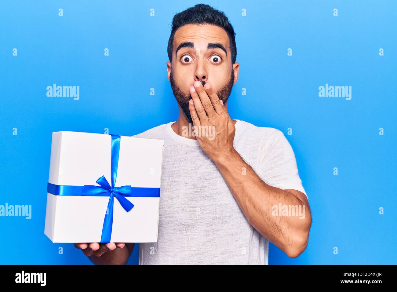 Young hispanic man holding gift covering mouth with hand, shocked and ...