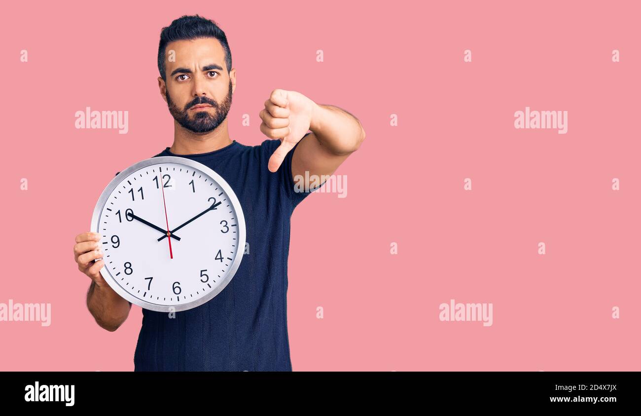 Young hispanic man holding big clock with angry face, negative sign ...