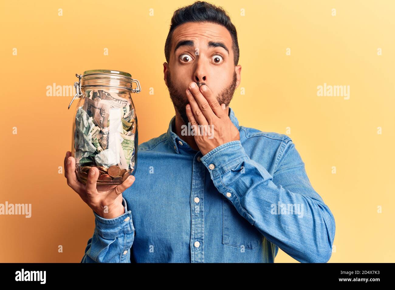 Young hispanic man holding jar with savings covering mouth with hand ...