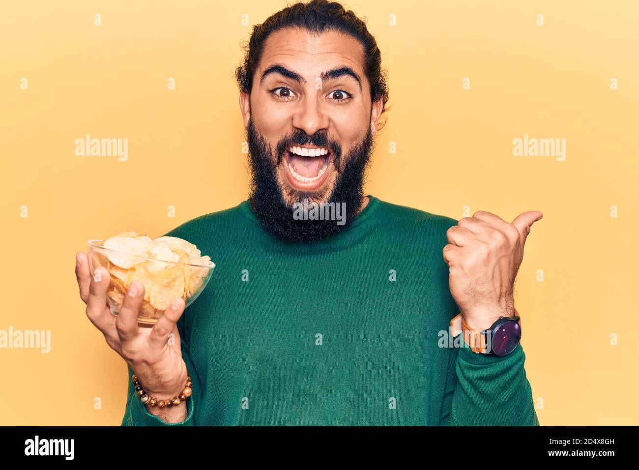 Young arab man holding potato chip pointing thumb up to the side ...