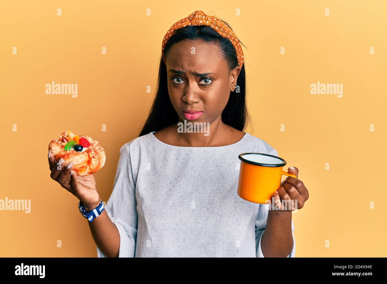 Young african woman drinking coffee and eating pastry clueless and ...