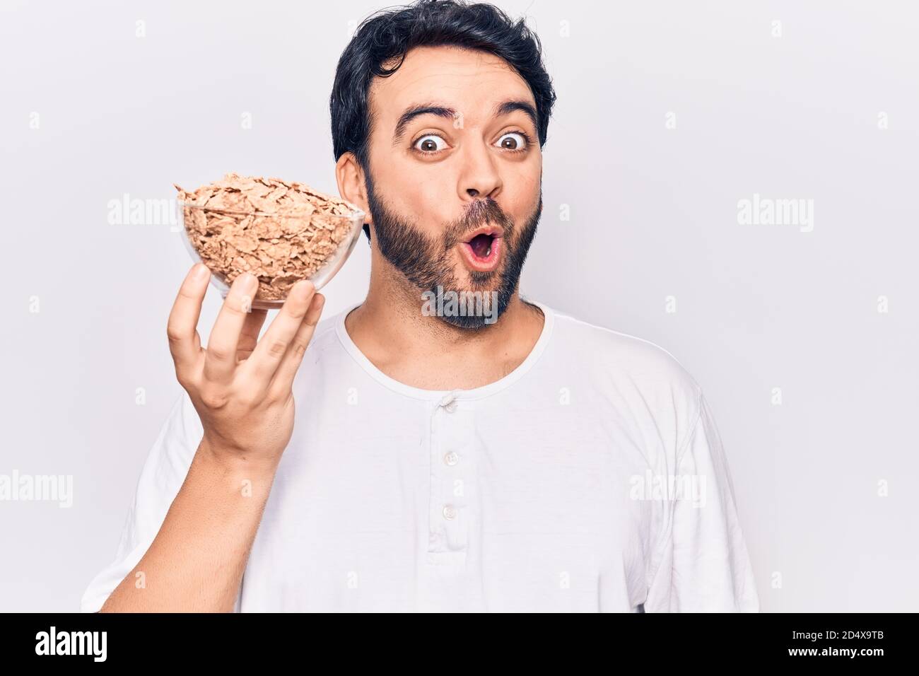 Young hispanic man holding cornflakes scared and amazed with open mouth ...