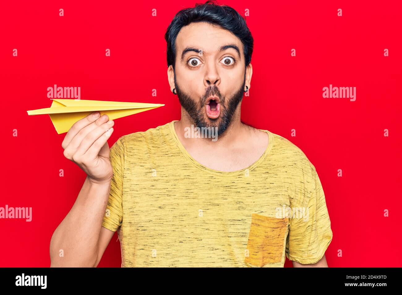 Young hispanic man holding paper airplane scared and amazed with open ...