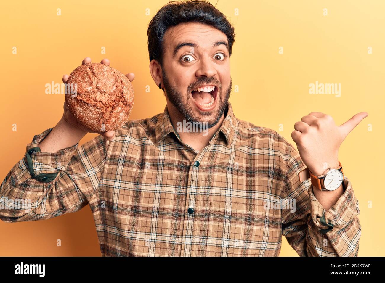 Young hispanic man holding bread pointing thumb up to the side smiling ...