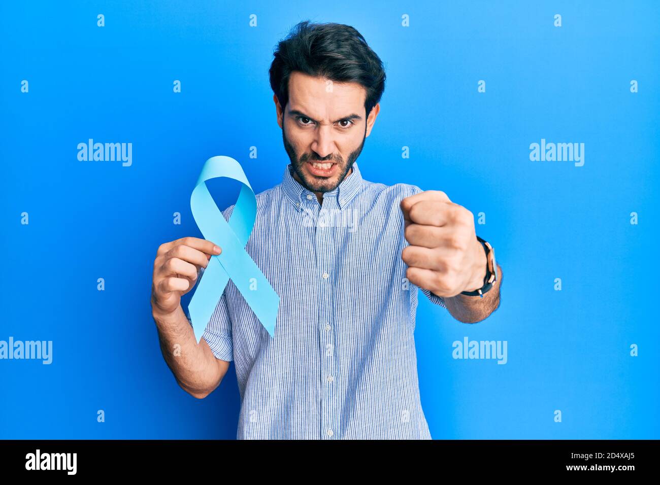 Young hispanic man holding blue ribbon annoyed and frustrated shouting ...