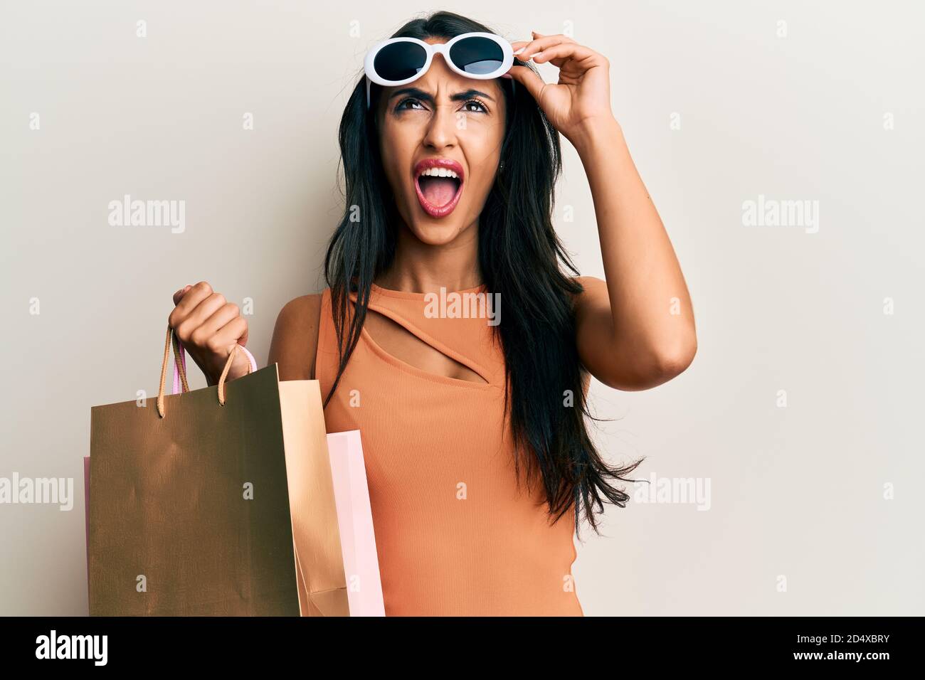 Beautiful hispanic woman holding shopping bags wearing sunglasses angry ...