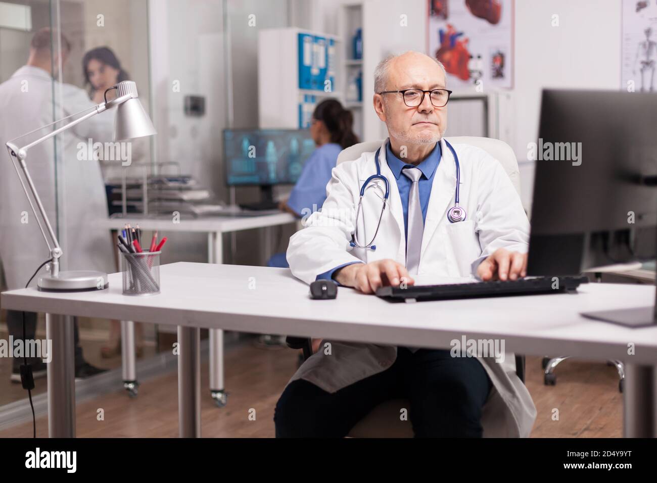 Senior doctor with stethoscope using pc in hospital cabinet while young ...