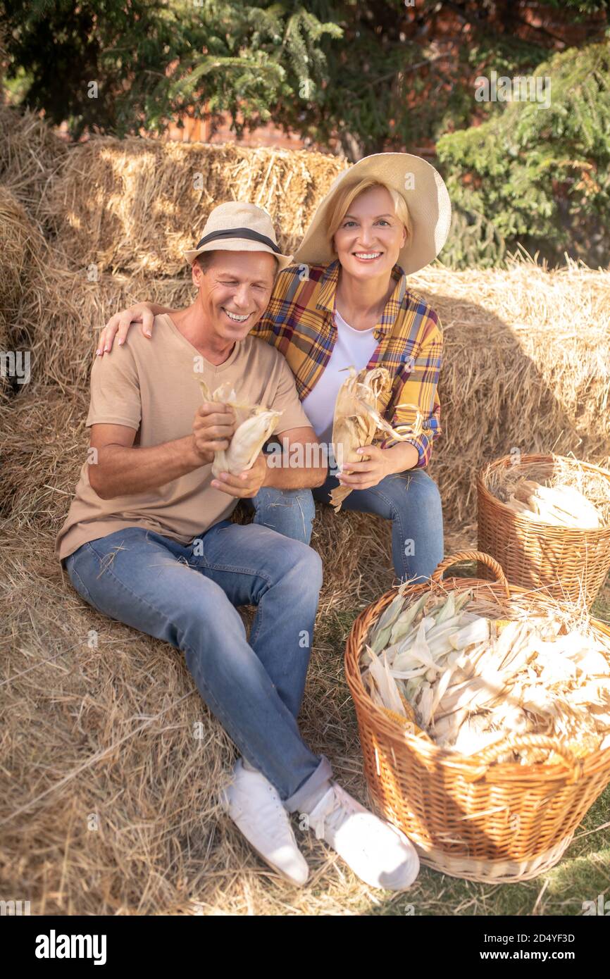 Male and female farmers sitting on hay, hugging, holding corn cobs ...