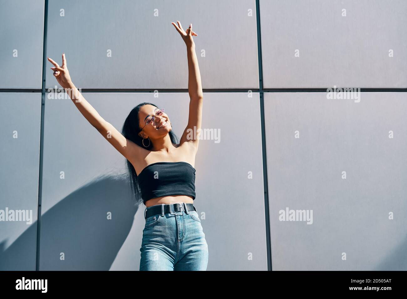 Fashionable cheerful young black woman with arms raised showing peace ...