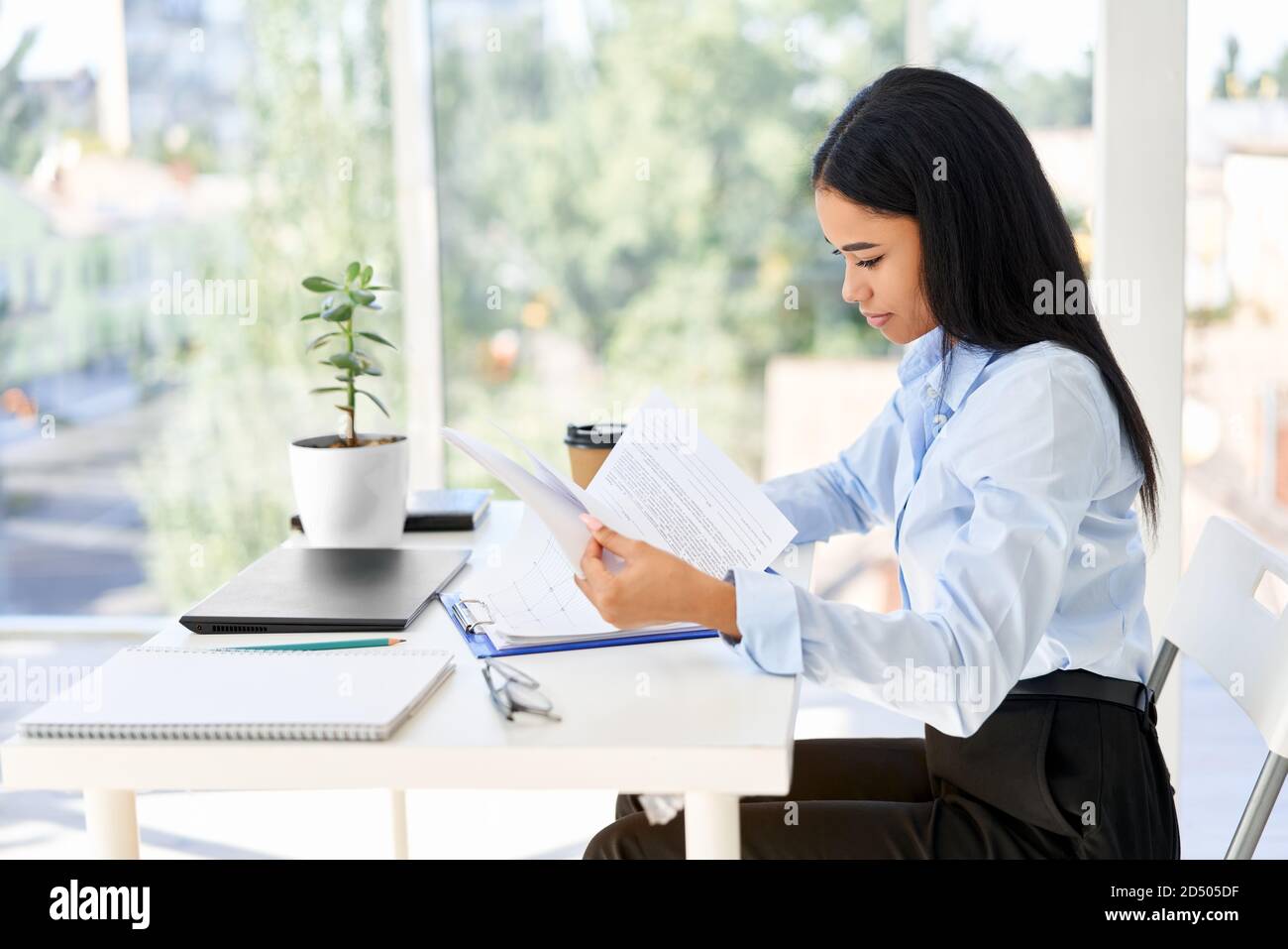 Pretty black businesswoman sitting at workplace and reading documents ...
