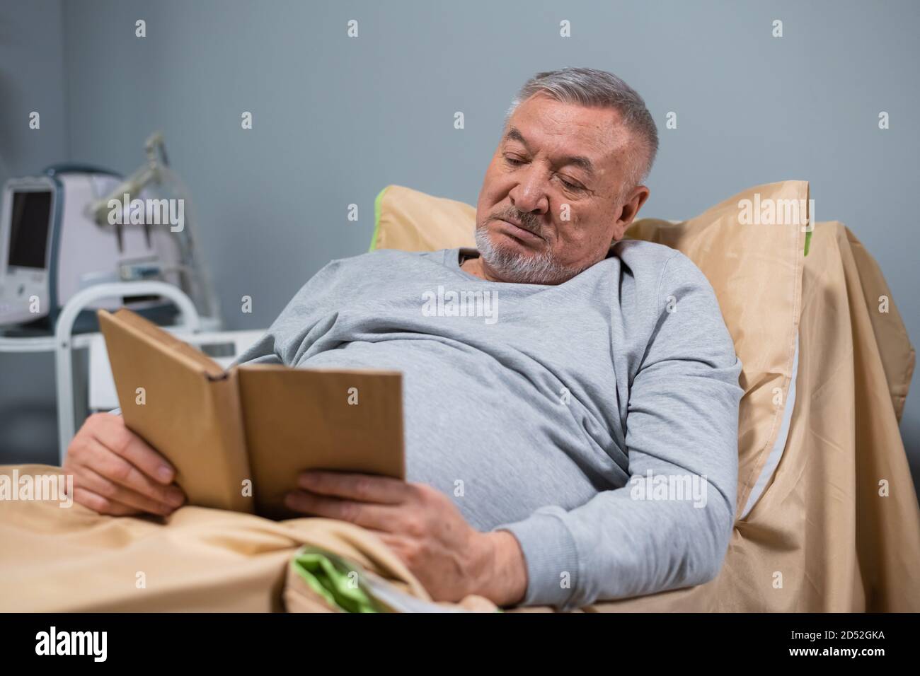 A gray-haired old man reading a book in his bed in a hospital ward ...
