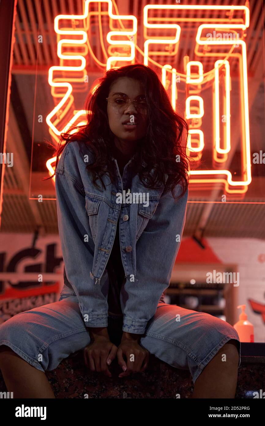 Hipster cool African gen z lady looking at camera sit under red neon ...
