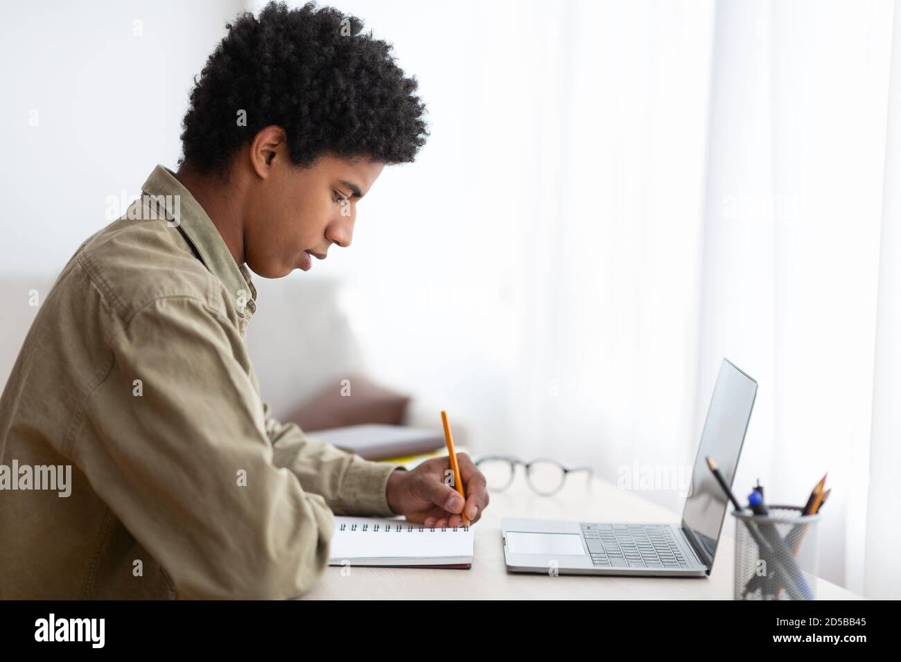 Remote education concept. Side view of black teen guy taking notes in ...
