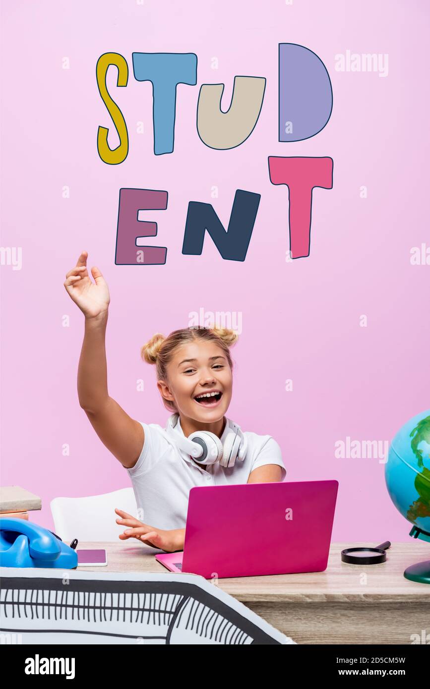 Schoolgirl sitting with raised hand near laptop, telephone, student ...