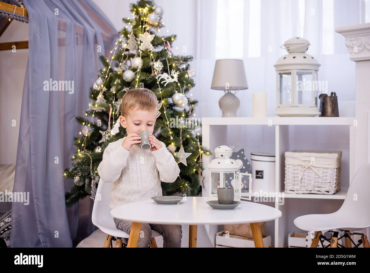 A little boy in a white pullover plays tea, drinks cocoa from a gray ...