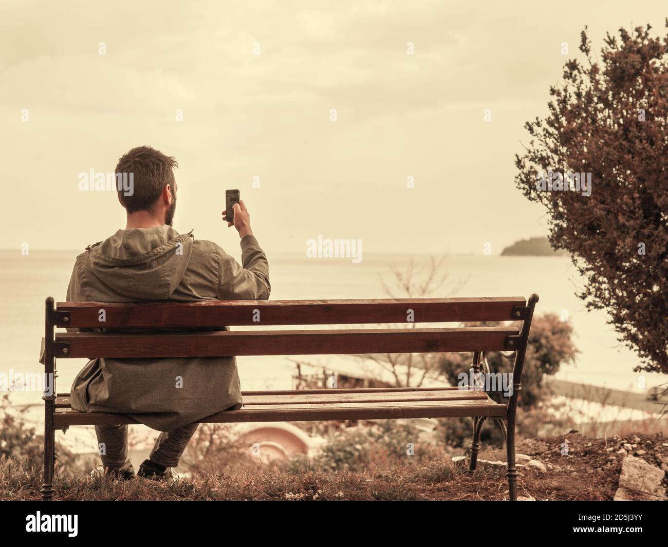 Man with back looking at phone screen while sitting on a bench outside ...