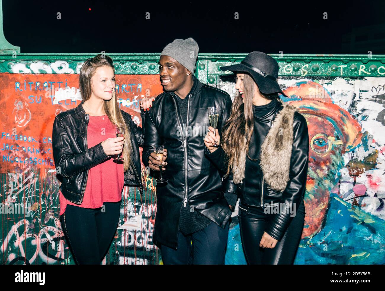 Friends holding champagne glasses standing at graffiti wall at night ...