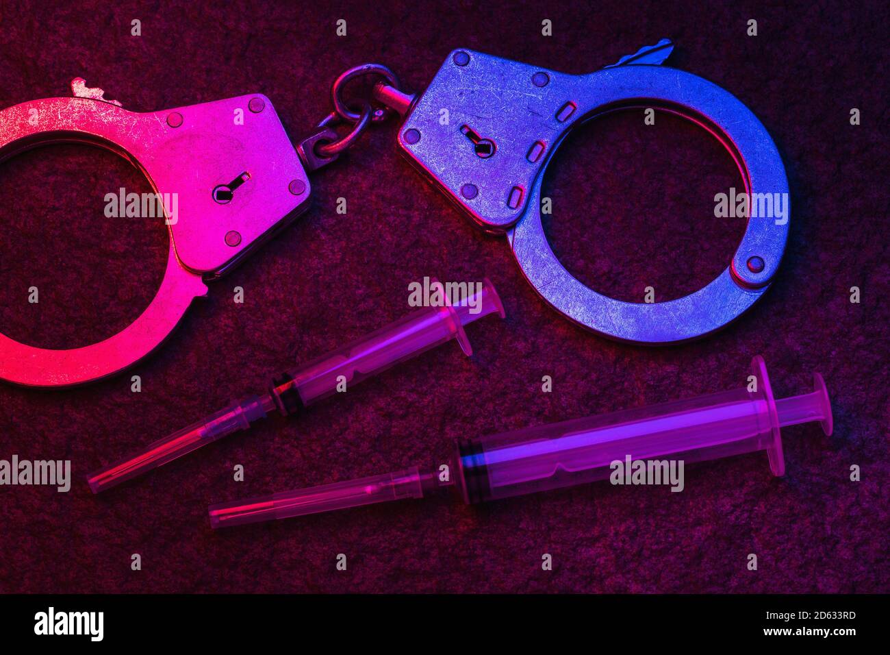 Handcuffs and syringes on a stone surface, lit by police car flashing ...