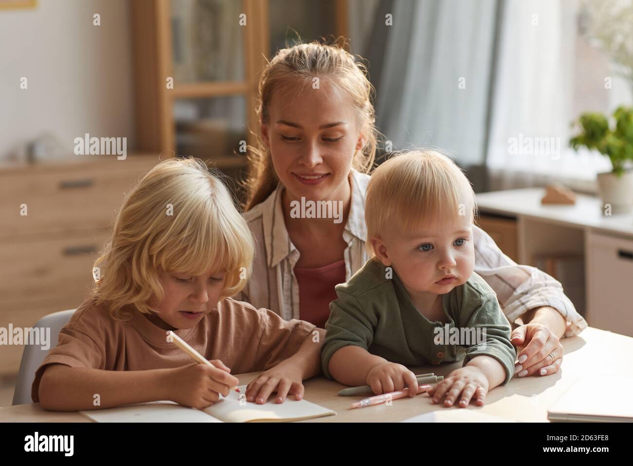 Young mother sitting at the table with baby and helping to her elder son with homework Stock ...