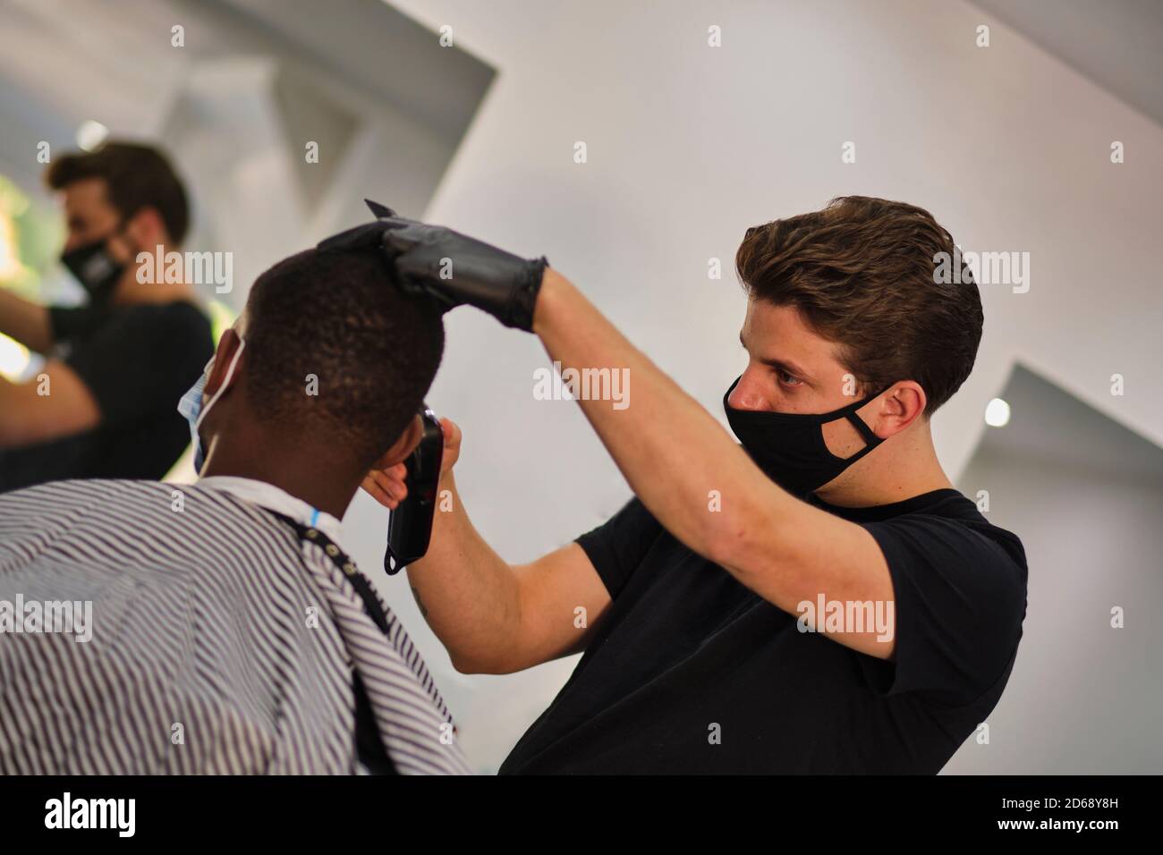 A young tattooed barber cuts a customers hair while wearing a face mask ...