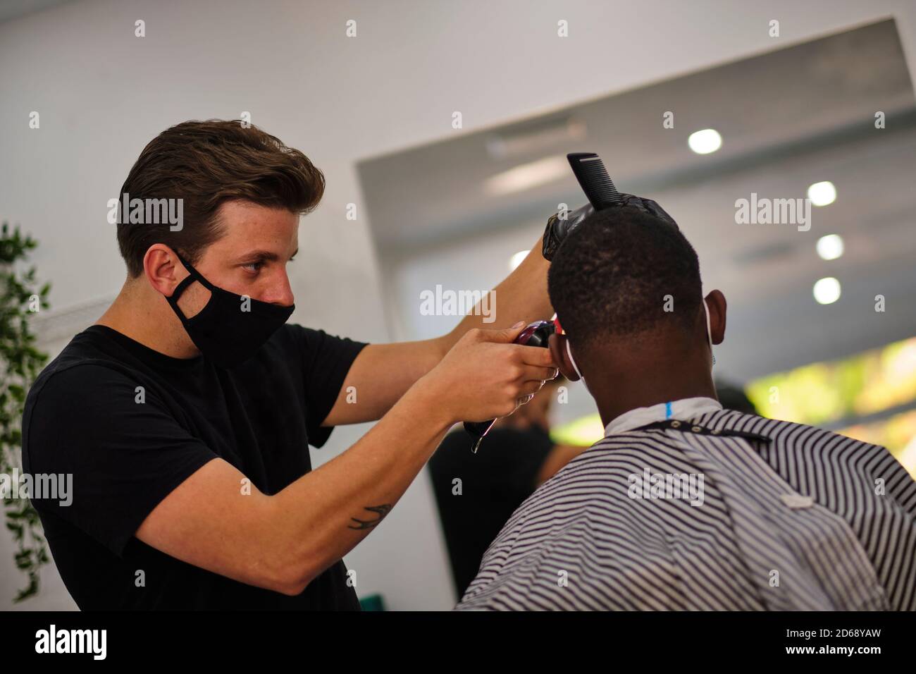 A young tattooed barber cuts a customers hair while wearing a face mask ...
